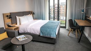 Quarto de hotel moderno com uma cama grande, uma mesa, uma cadeira e vista para edifícios pela janela.