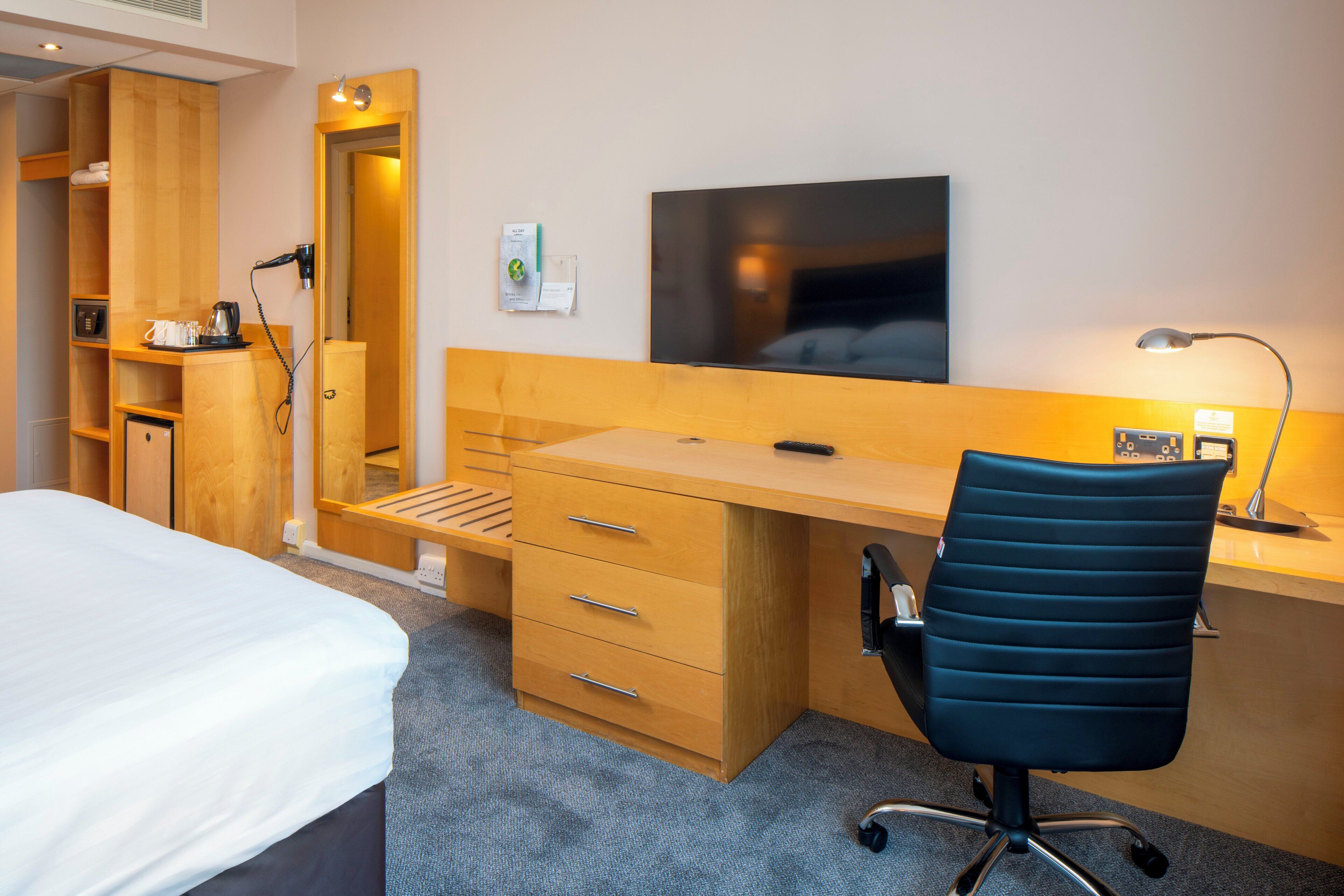 Quarto de hotel com cama, escrivaninha de madeira, cadeira, televisão e pequeno armário com chaleira.