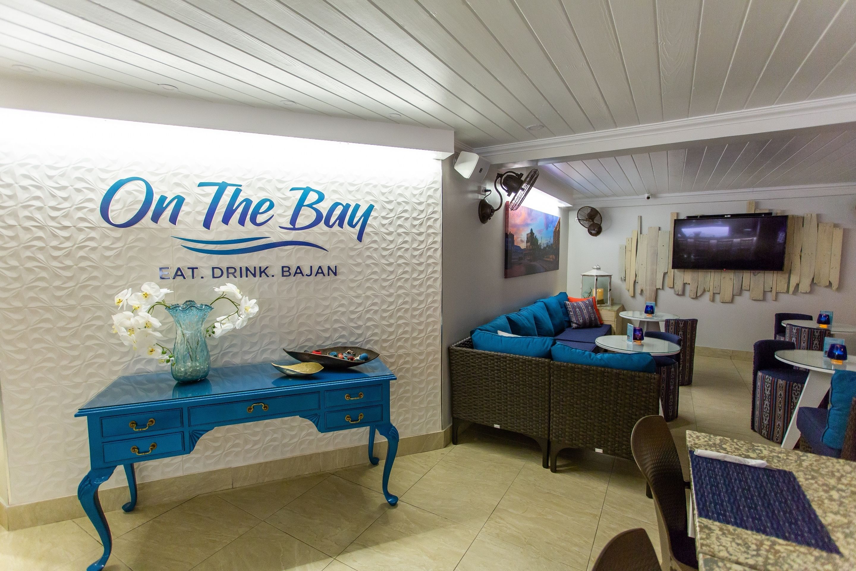 Quarto com escrivaninha azul, mobiliário de vime, televisão na parede e uma placa com a inscrição "On The Bay".