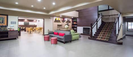 Um hall de hotel espaçoso com uma escadaria moderna, uma área de estar com sofá e poltronas, e uma área de jantar ao fundo.