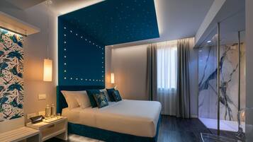 Quarto de hotel moderno com uma cama grande, mesa de cabeceira, escrivaninha e chuveiro em cabine de vidro.