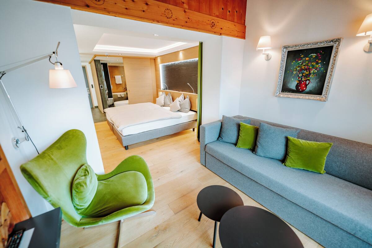 Quarto de hotel moderno com uma cama, um sofá cinza com almofadas verdes, uma poltrona verde e uma pequena mesa.