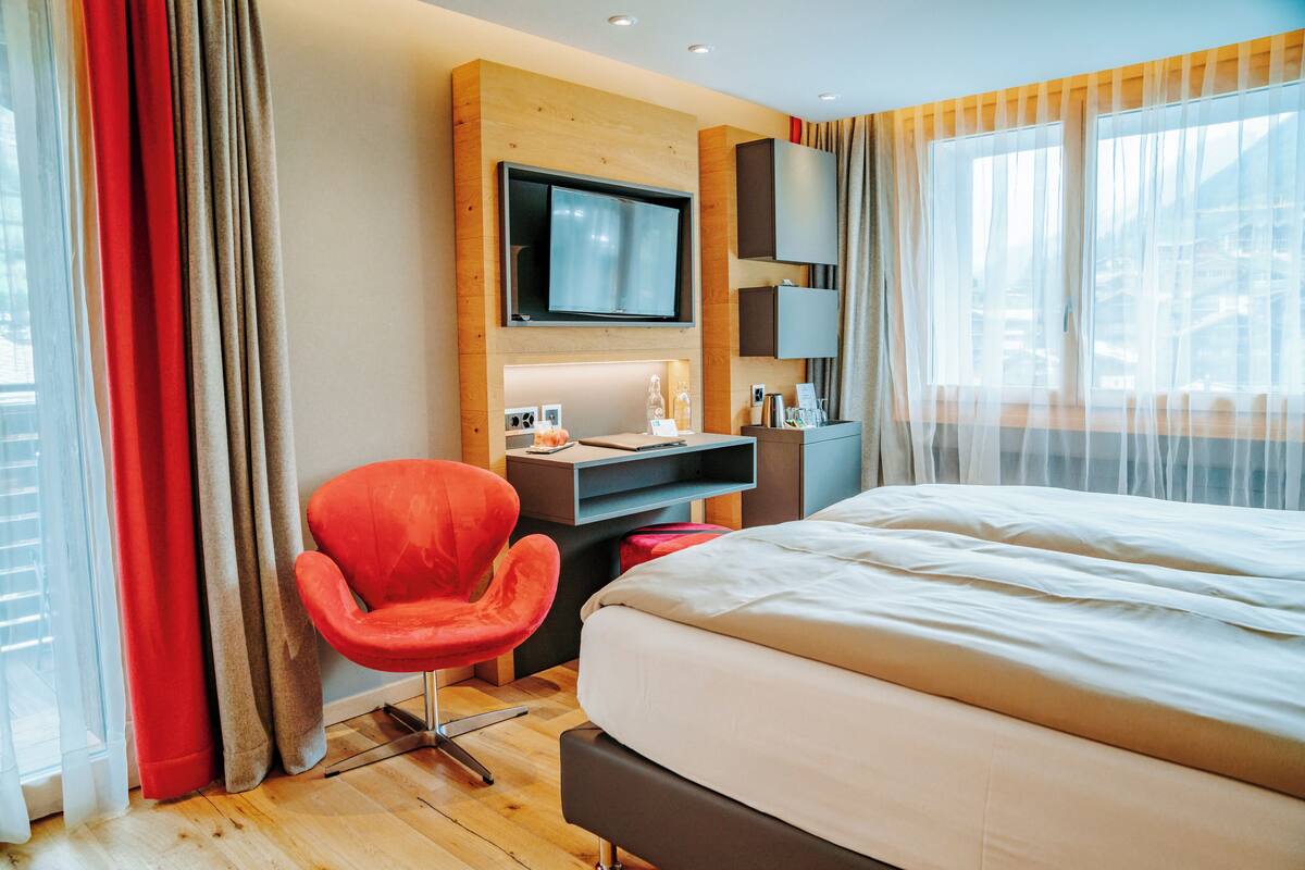 Quarto de hotel moderno com uma cama grande, uma escrivaninha com cadeira, uma TV de tela plana e uma janela ampla com cortinas leves.