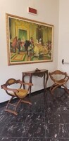 Quarto com uma pintura grande na parede, duas cadeiras de madeira e uma mesinha.
