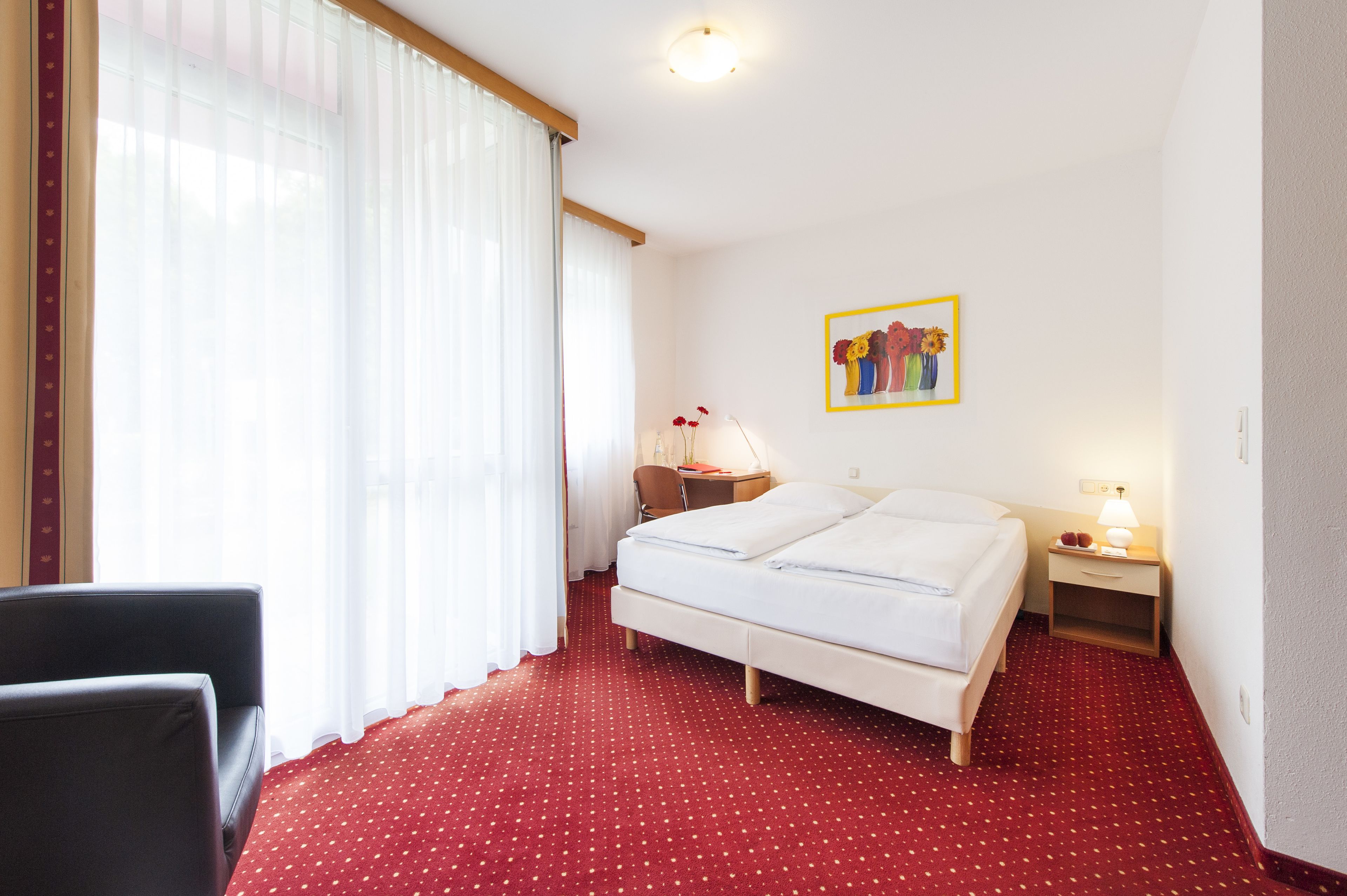 Quarto de hotel com uma cama, uma escrivaninha com candeeiro, uma cadeira e um quadro na parede.