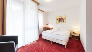 Quarto de hotel com uma cama, uma escrivaninha com candeeiro, uma cadeira e um quadro na parede.