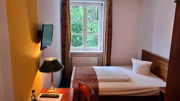 Quarto de hotel com uma cama, uma escrivaninha com abajur, uma televisão na parede e uma janela com cortinas.
