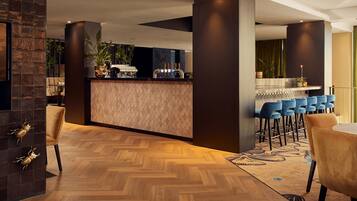 Uma área de bar moderna com piso de madeira em padrão herringbone, um balcão central e banquetas azuis.