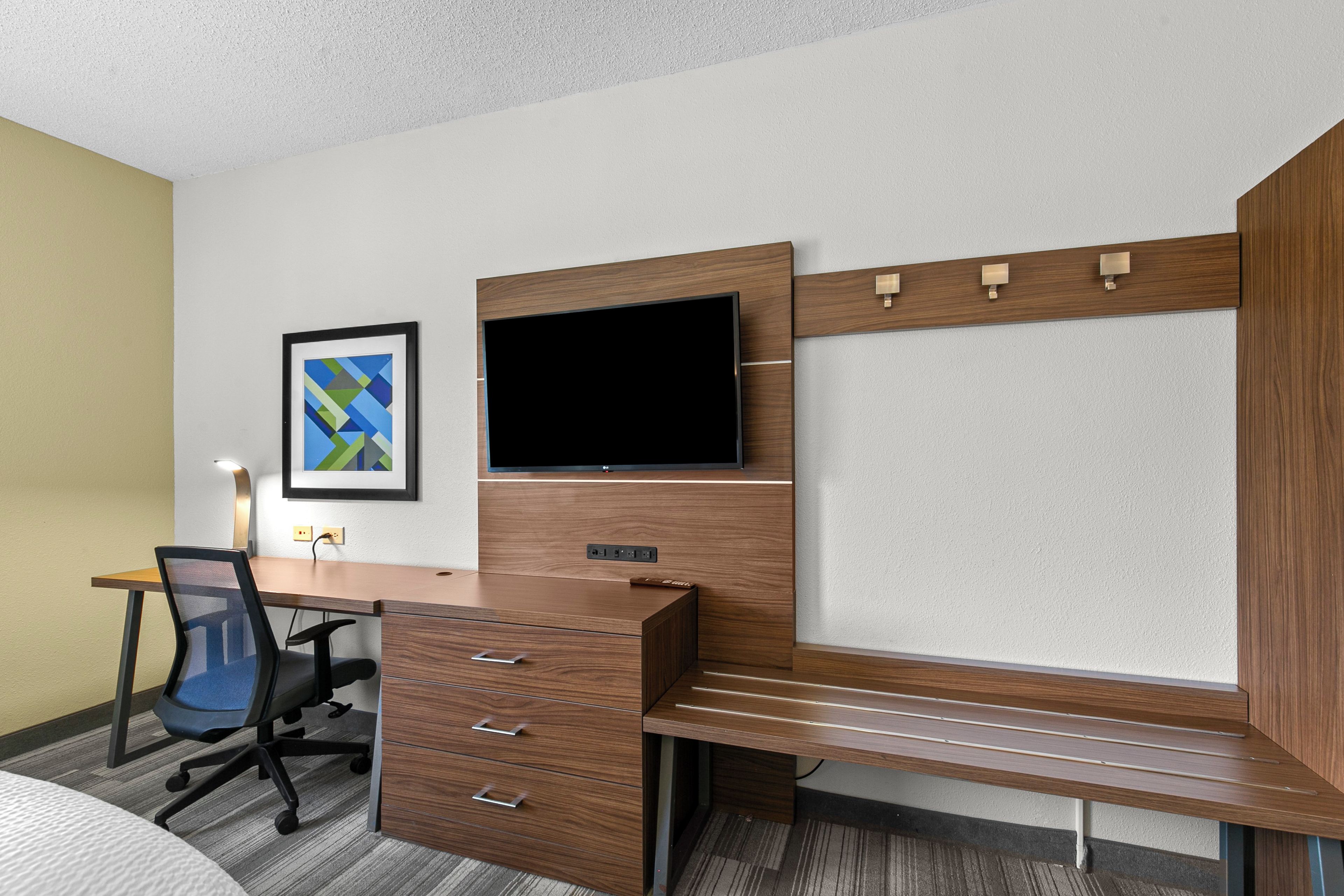 Quarto de hotel com escrivaninha de madeira, televisão de tela plana, quadro pendurado na parede e uma cama.