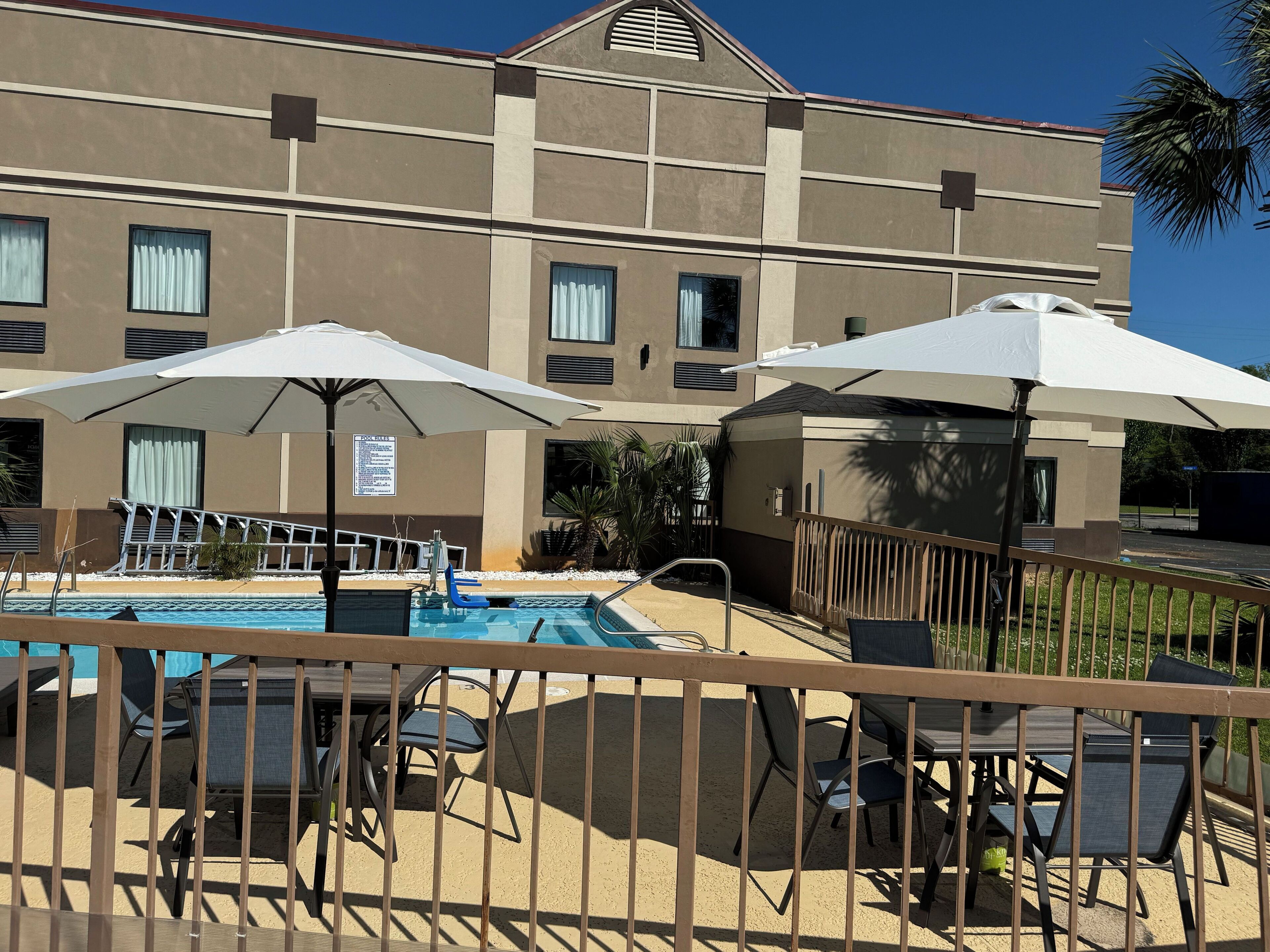Área de piscina com espreguiçadeiras e guarda-sóis, um edifício com várias janelas, e céu claro.