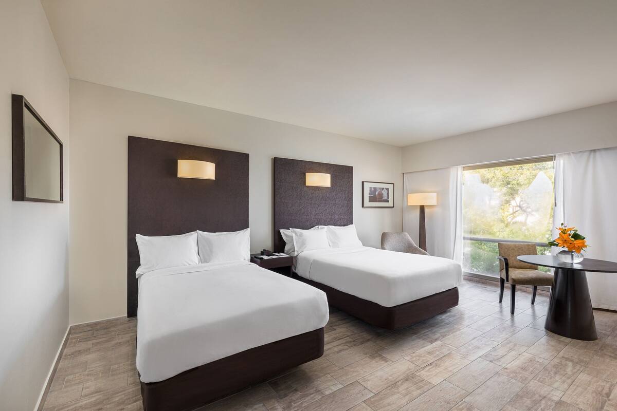 Quarto de hotel com duas camas, uma mesinha, uma cadeira e janela com vista para árvores.
