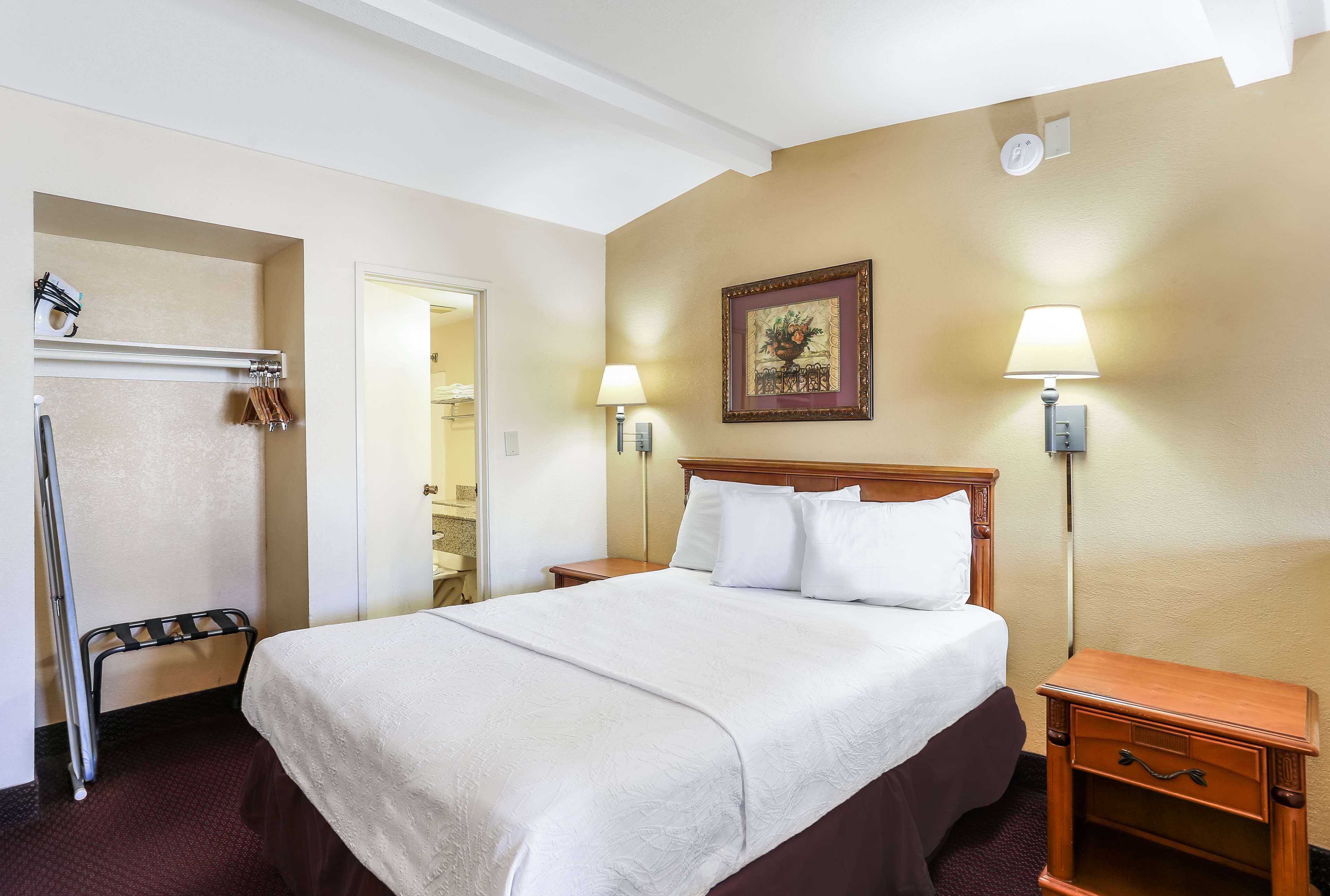 Quarto de hotel com uma cama grande, uma mesinha de cabeceira de madeira, um abajur, um cabideiro e uma porta que dá para um banheiro.