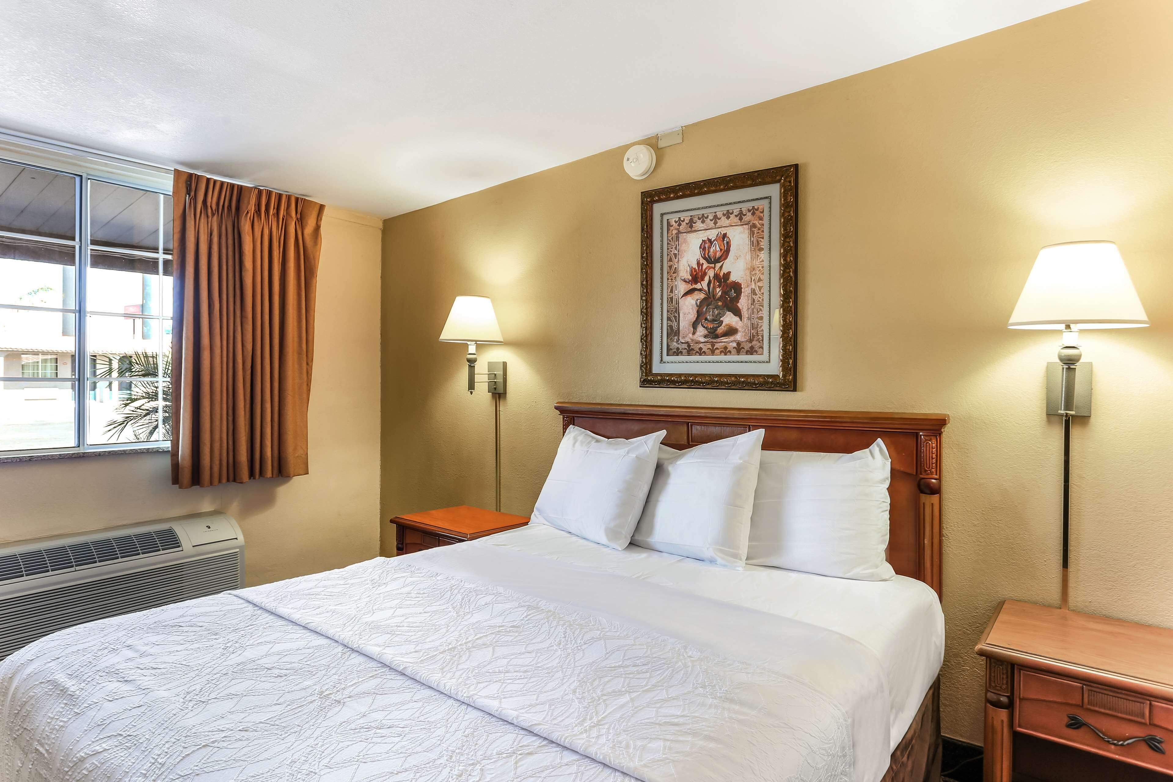 Quarto de hotel com uma cama grande, duas luminárias de cabeceira, uma pintura floral emoldurada na parede e uma janela com cortinas.