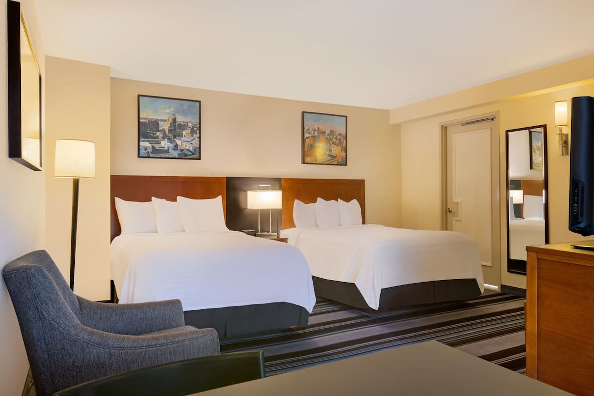 Quarto de hotel com duas camas, uma escrivaninha, uma cadeira, televisão e obras de arte nas paredes.