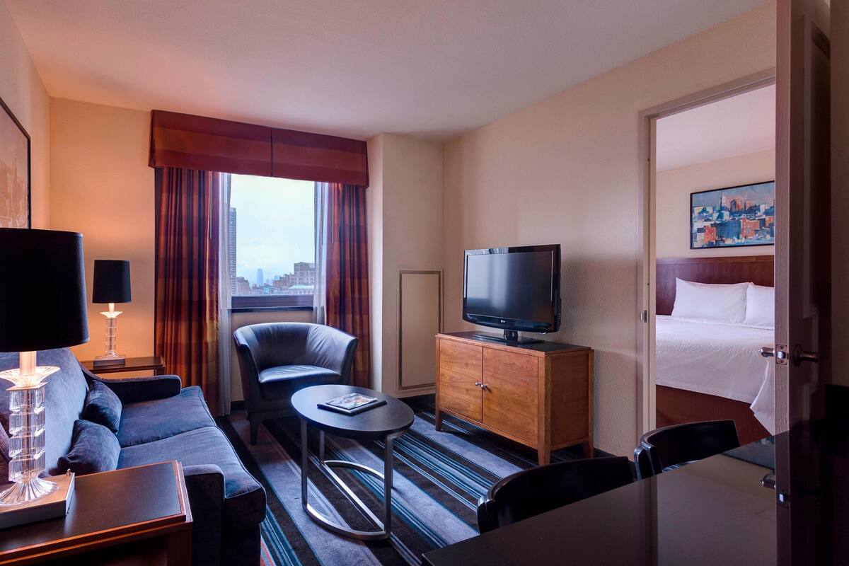 Quarto de hotel com sofá, uma pequena mesa, televisão sobre um móvel de madeira, cama com roupa de cama branca e vista para a cidade pela janela.