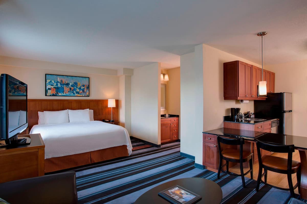 Quarto de hotel com cama, escrivaninha com cadeira, televisão, pintura na parede e área de cozinha com armários e frigorífico.