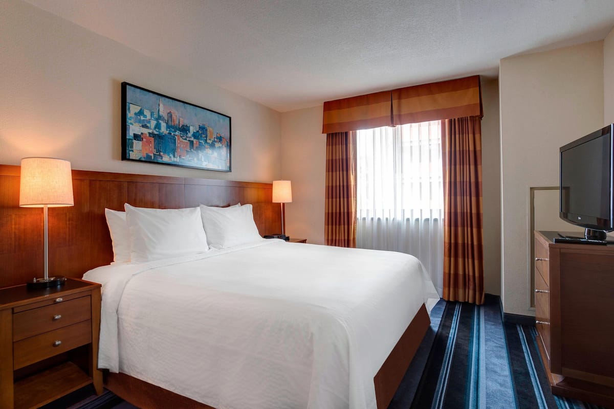 Quarto de hotel com uma cama grande, duas mesas de cabeceira com luminárias, uma televisão sobre um armário de madeira e um quadro na parede.