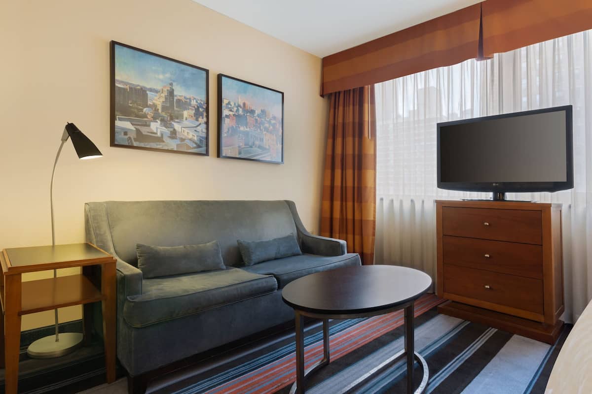 Quarto de hotel com sofá, uma mesinha, televisão e duas pinturas na parede.