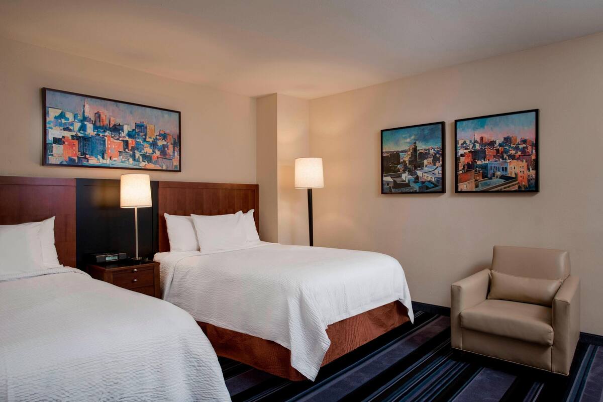 Quarto de hotel com duas camas, uma cadeira e duas pinturas de paisagens urbanas nas paredes.