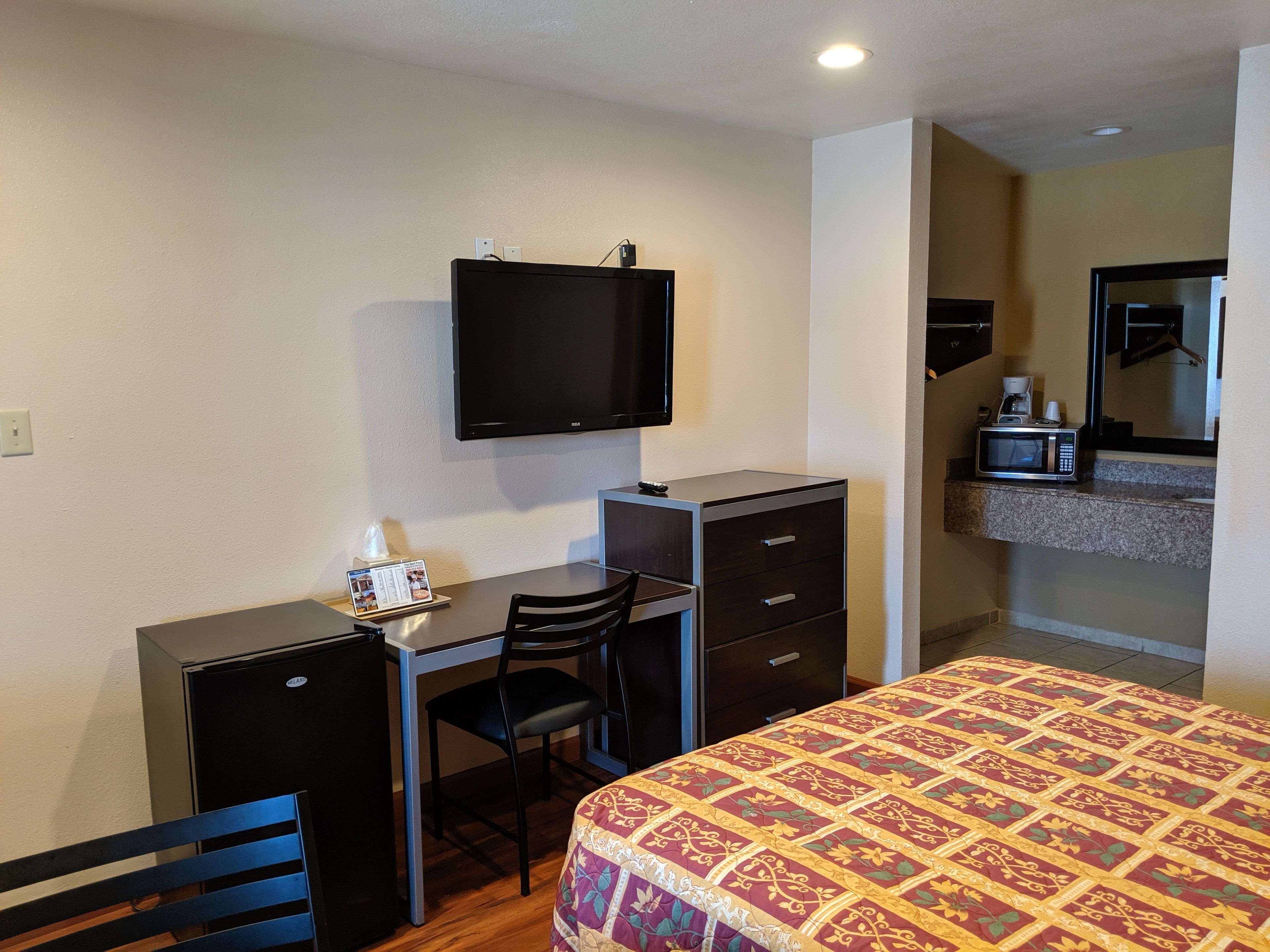 Quarto de hotel compacto com cama, escrivaninha com cadeira, televisão, pequena geladeira, micro-ondas e cafeteira.