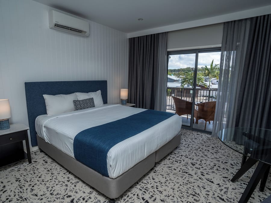 Quarto de hotel com uma cama grande, mesa de cabeceira com abajur, mesinha de vidro e vista para o exterior por uma porta de correr.