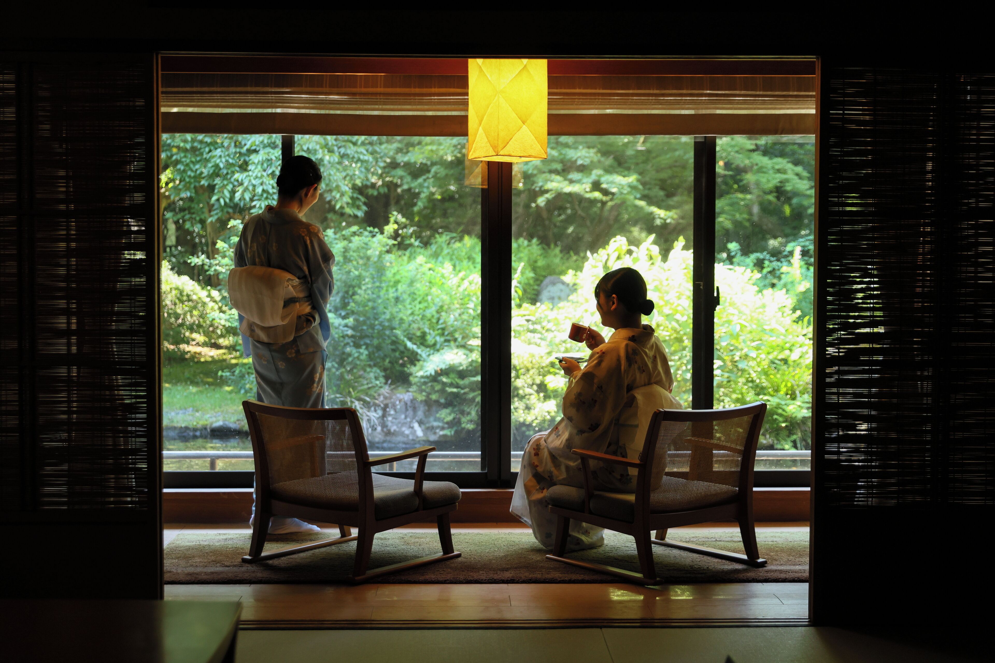 Duas pessoas sentadas perto de uma janela com vista para o verde, uma luminária pendente e mobiliário tradicional.