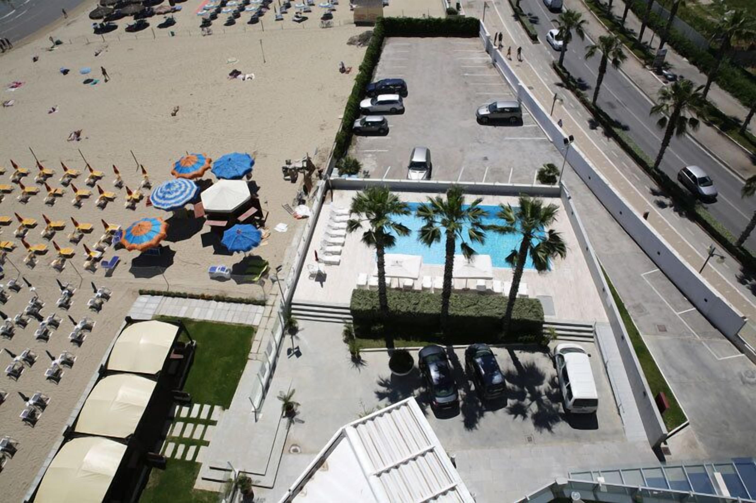Vista aérea de uma praia com guarda-sóis, espreguiçadeiras, piscina e carros estacionados.