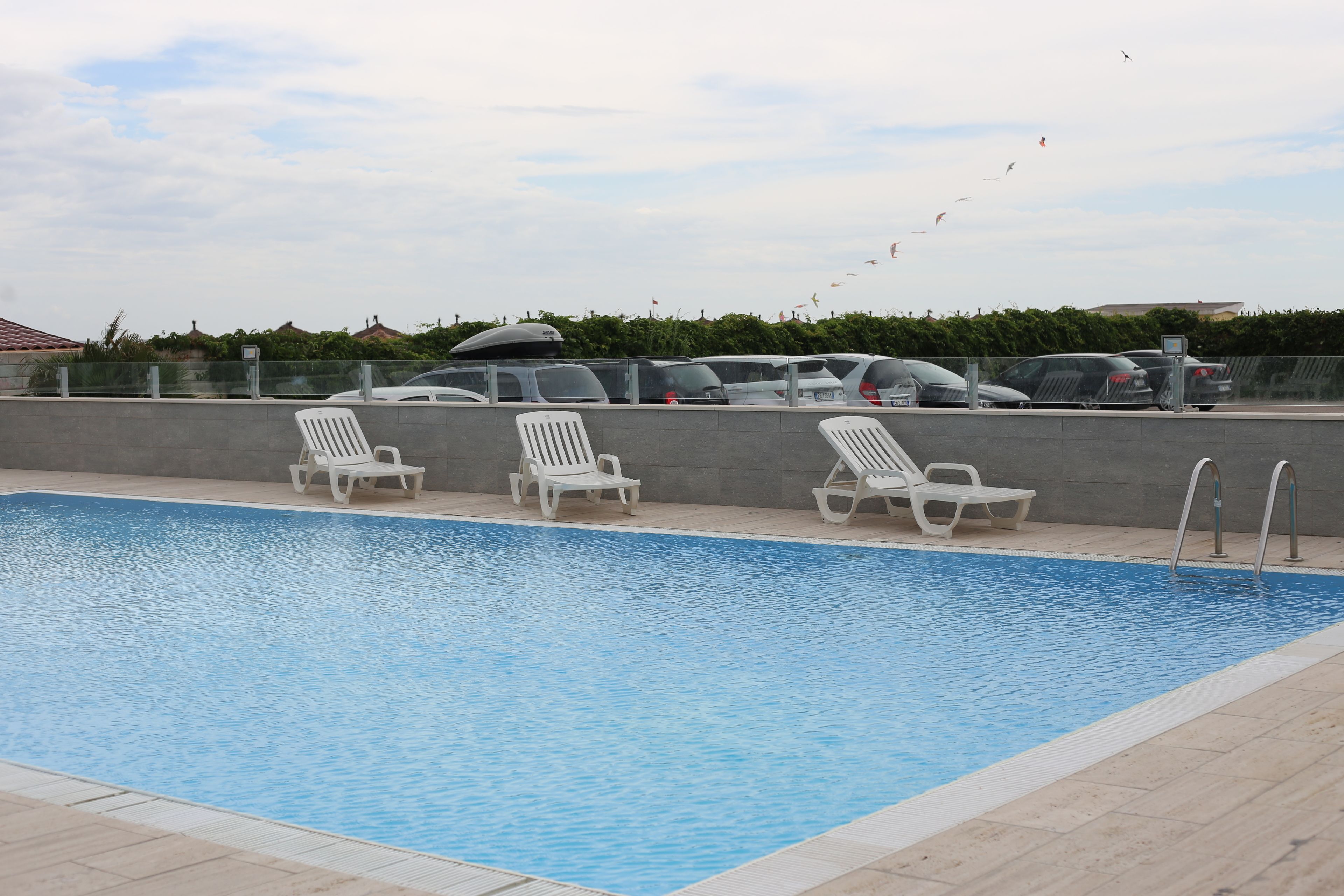 Piscina com três espreguiçadeiras brancas e uma fileira de carros estacionados ao fundo.