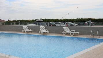 Piscina com três espreguiçadeiras brancas e uma fileira de carros estacionados ao fundo.