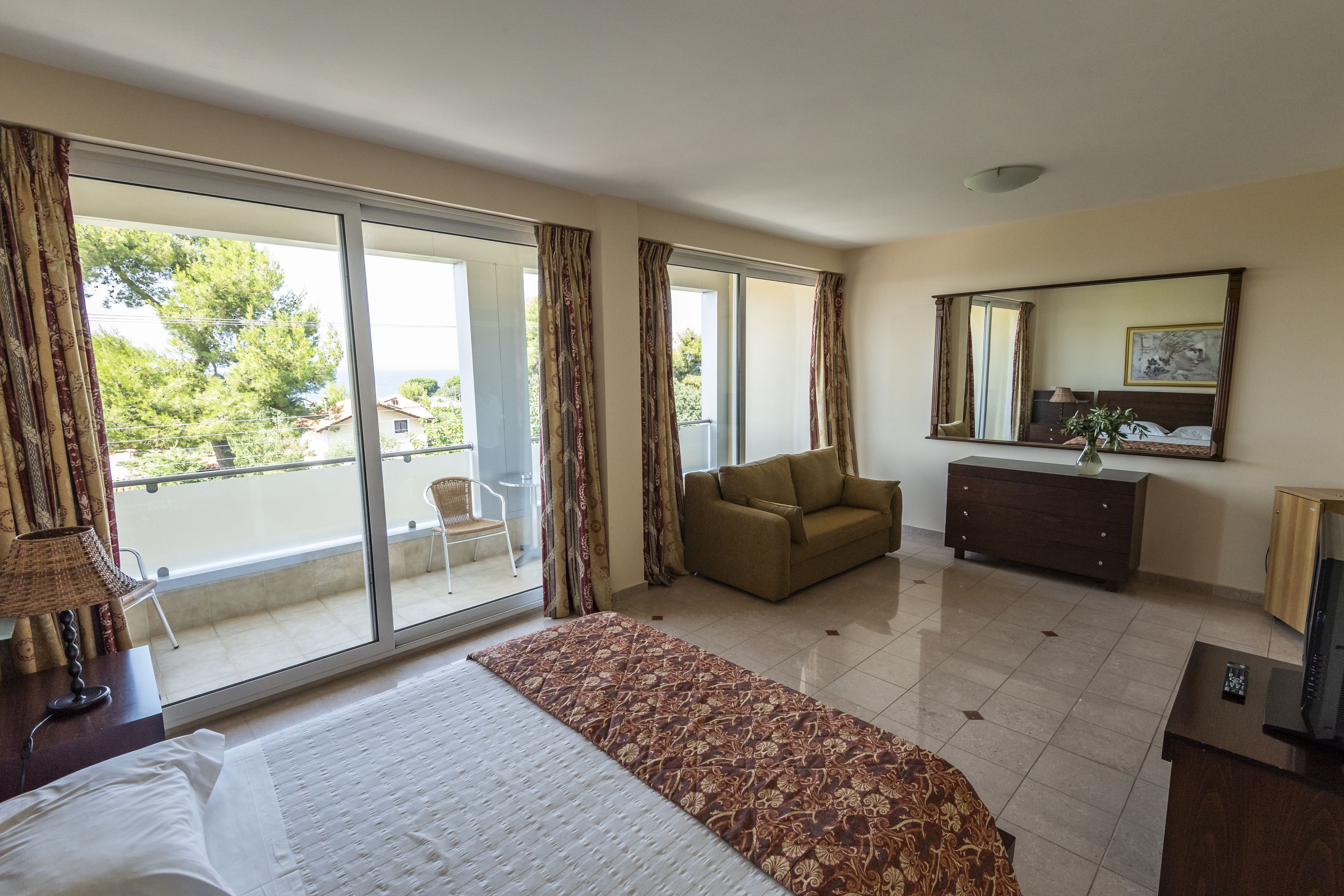 Quarto de hotel com uma cama grande, um sofá, um aparador, um espelho e uma varanda com vista para árvores e edifícios.