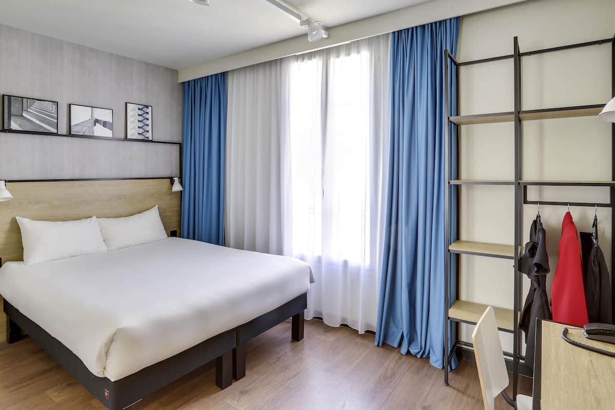 Quarto de hotel com uma cama, uma escrivaninha com cadeira, uma estante com roupas e uma janela com cortinas.