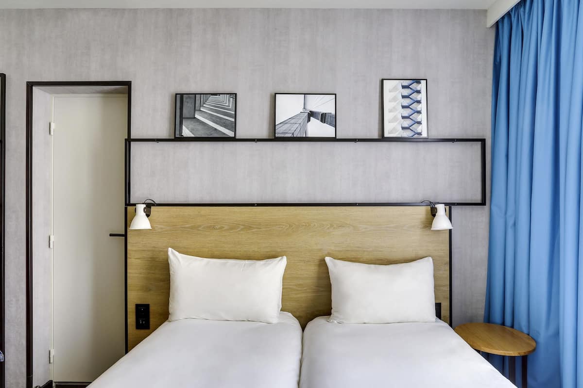 Quarto de hotel com duas camas, cabeceira de madeira, uma mesinha de apoio e prateleira fixada na parede com quadros emoldurados.