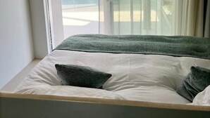 Quarto com uma janela grande, uma cama com roupa de cama branca e um cobertor verde.