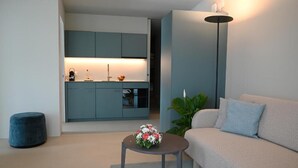 Uma sala moderna com um sofá cinzento, uma mesinha redonda com um arranjo floral e uma área de cozinha com armários verde-água e uma ilha iluminada.