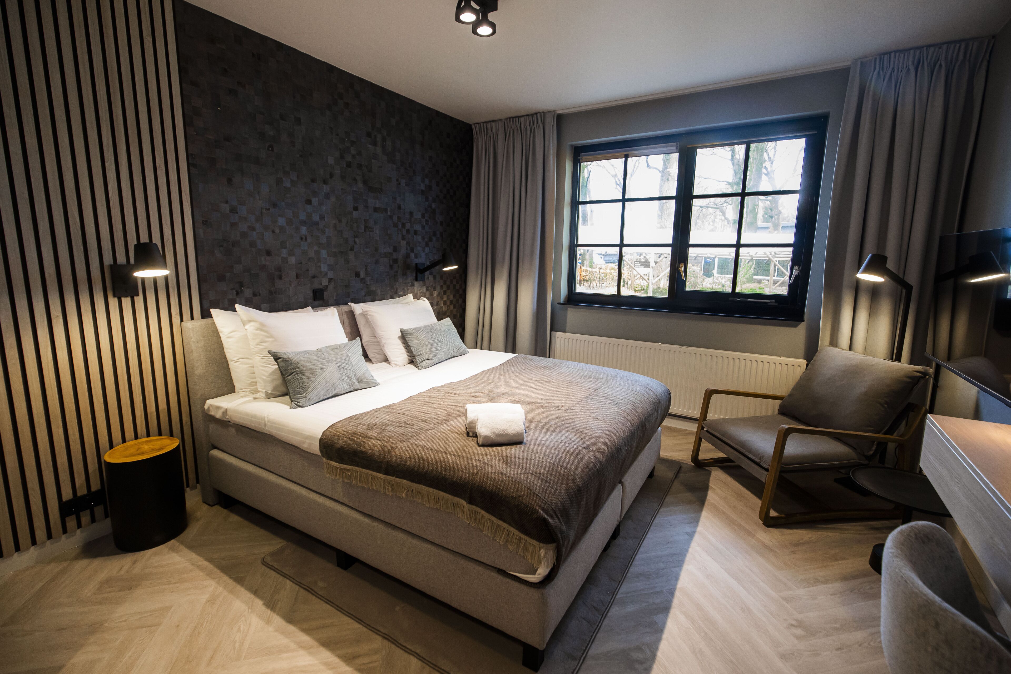 Quarto moderno com uma cama grande, uma janela com cortinas, uma mesinha de cabeceira e uma cadeira.