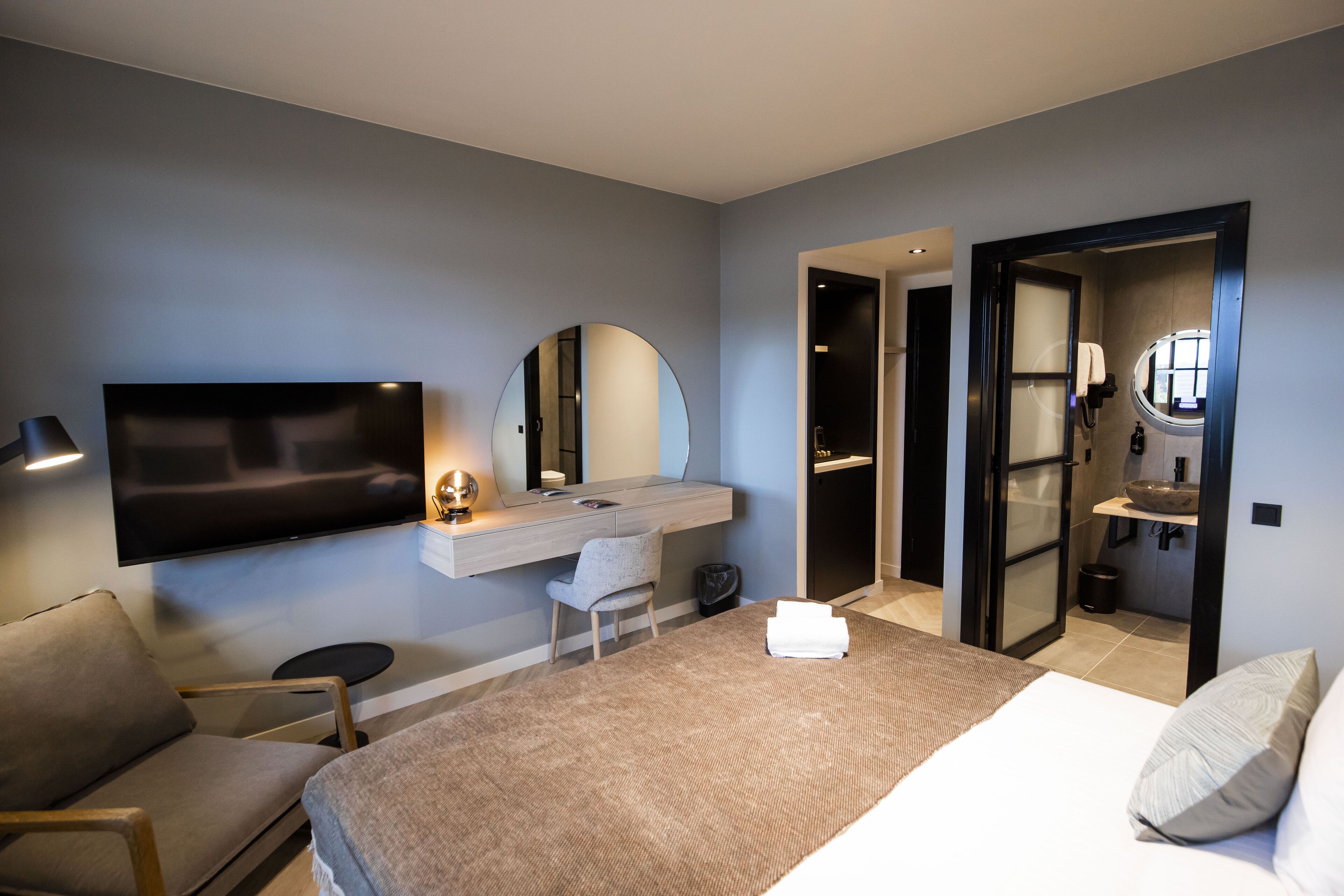 Quarto de hotel moderno com uma cama grande, uma escrivaninha com espelho, televisão de tela plana, sofá e um banheiro visível através de uma porta aberta.