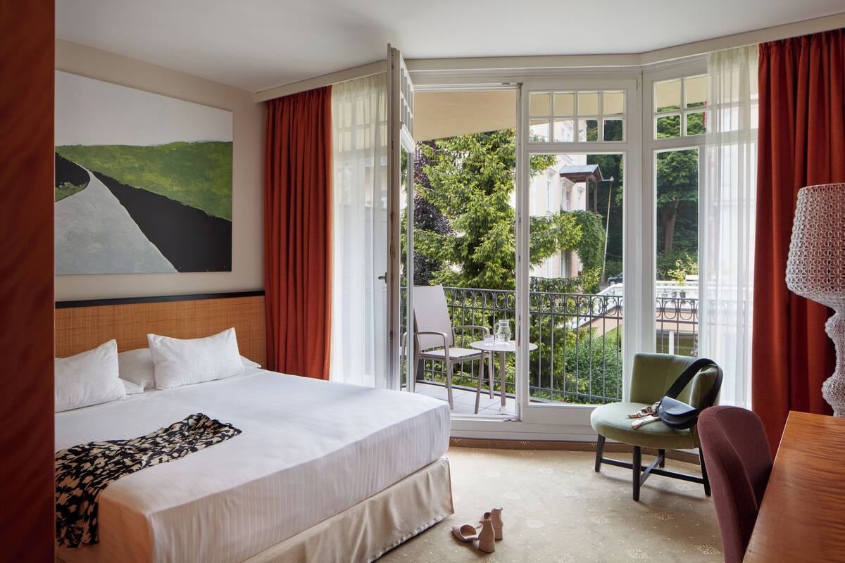 Quarto de hotel com uma cama grande, um quadro na parede, uma varanda com cadeira e mesa, e vista para o verde lá fora.