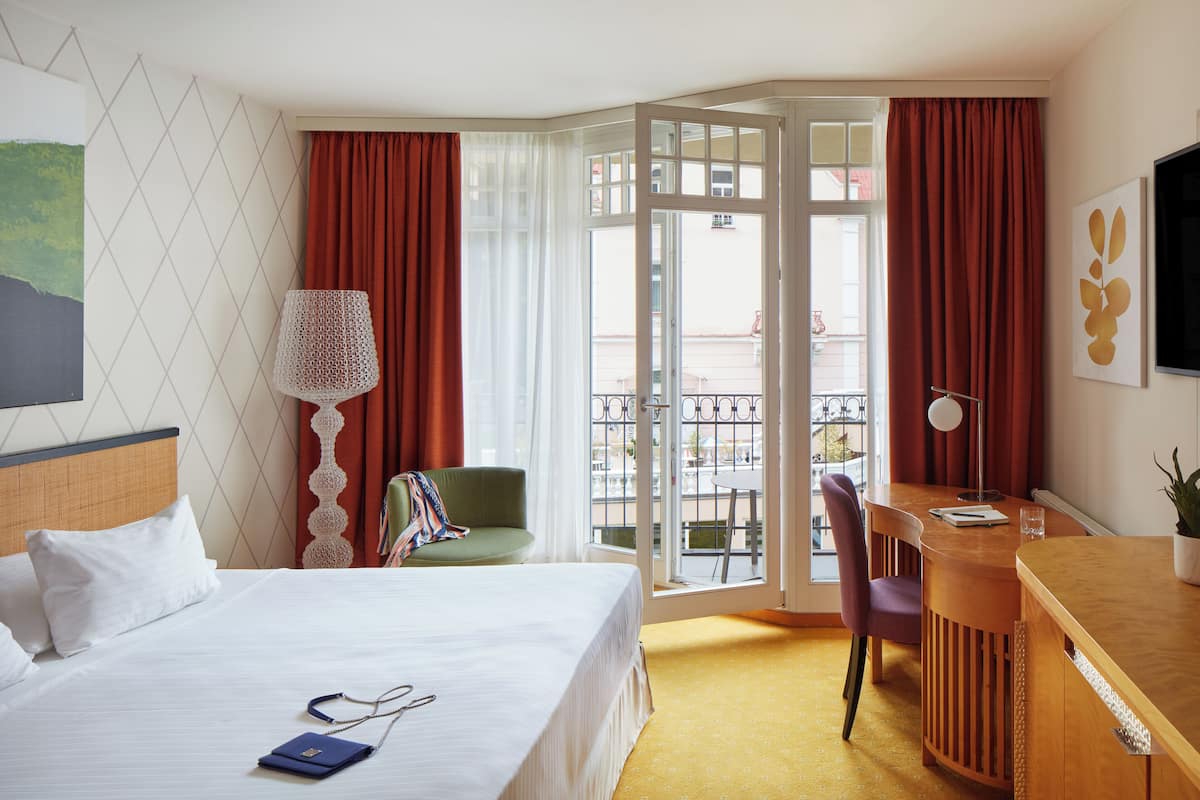 Quarto de hotel com cama, escrivaninha, cadeira, luminária e varanda com cortinas.