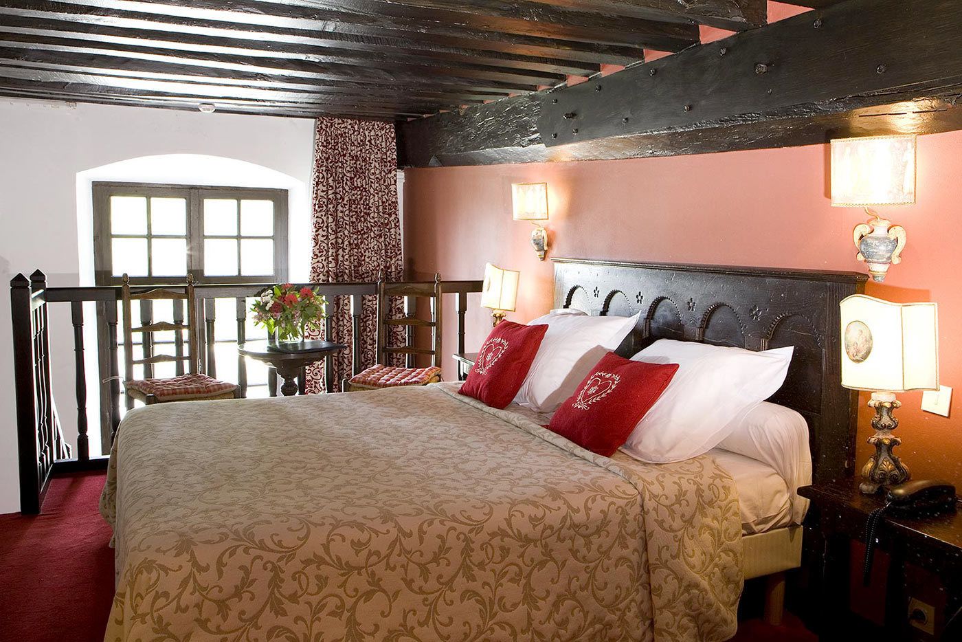 Quarto com cama grande, cabeceira de madeira, duas luminárias de cabeceira, mesa de cabeceira, pequena mesa com um vaso de flores, uma cadeira e varanda com grade.