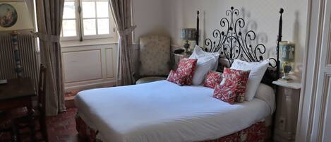 Uma cama bem arrumada com colcha floral e lençóis brancos, uma cadeira de madeira, um radiador e uma janela com cortinas.