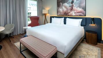 Quarto de hotel com uma cama grande, uma cadeira vermelha, uma mesa pequena, um banco, uma luminária e um quadro na parede.