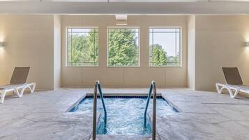 Piscina interna com vista clara para árvores através de grandes janelas, rodeada por cadeiras de descanso brancas.