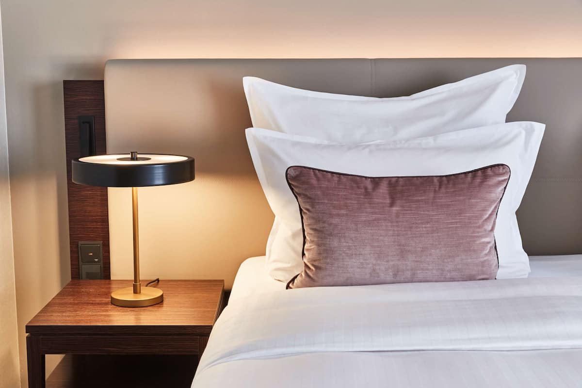 Uma cama bem arrumada com almofadas brancas e castanhas, uma luminária de cabeceira e uma mesa de cabeceira de madeira.