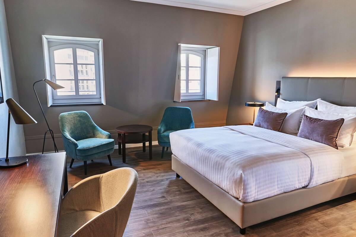 Quarto de hotel com uma cama grande, duas poltronas, uma mesinha e duas janelas.
