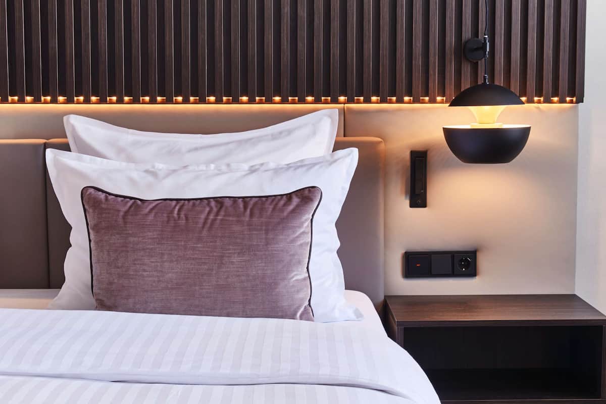 Cama bem arrumada com almofadas brancas e castanhas, mesa de cabeceira escura com abajur preto, e parede com ripas de madeira vertical e fileira de luzes.