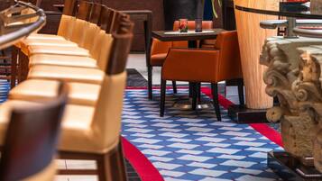 Um saguão de hotel com piso xadrez, uma fileira de cadeiras de madeira, uma mesa com cadeiras laranja, um pilar decorativo e uma porta com a inscrição “FESTSAAL”.
