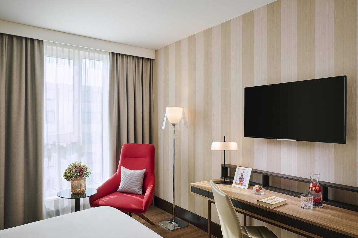 Quarto de hotel com cama, poltrona vermelha, escrivaninha com luminária, TV de tela plana e janela com cortinas.