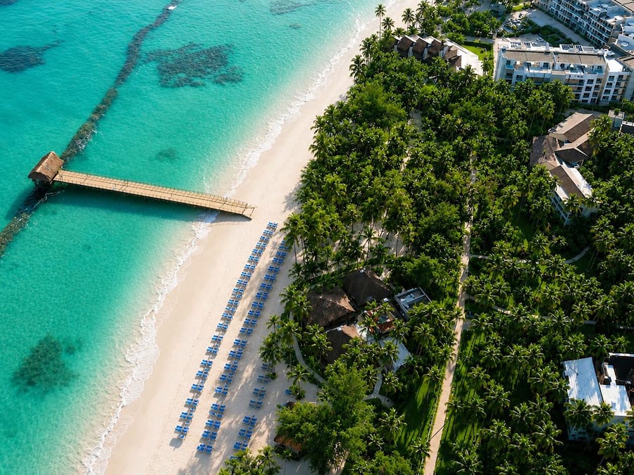 Vista aérea de um resort à beira-mar, com um píer, fileiras de cadeiras de praia e palmeiras exuberantes.