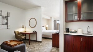 Quarto de hotel com cama, escrivaninha com luminária, cadeira, mesinha com bandeja e área de cozinha compacta com pia e armários.