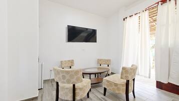 Quarto com televisão de tela plana na parede, uma mesa redonda com quatro cadeiras estampadas e uma janela com cortinas leves.